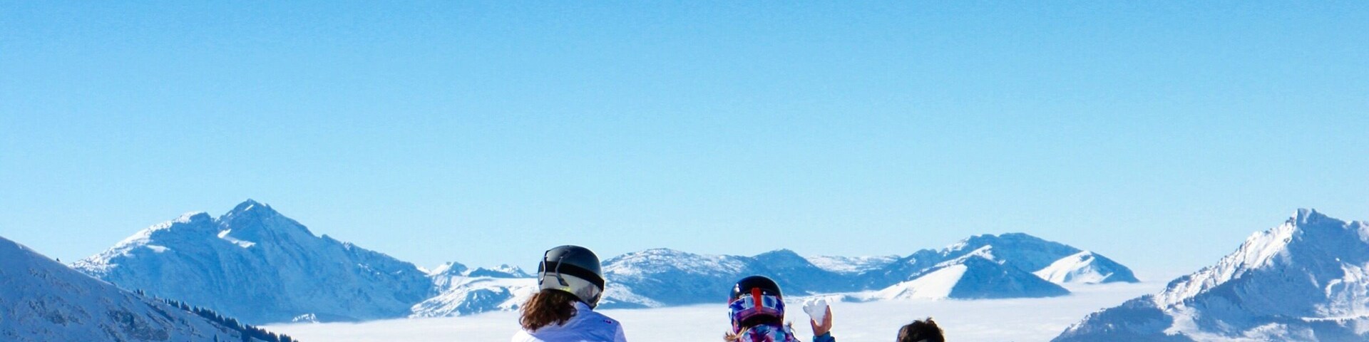 Paisagem nevada de montanha com três pessoas sentadas numa encosta coberta de neve, observando uma cadeia de picos cobertos de neve sob um céu azul claro.