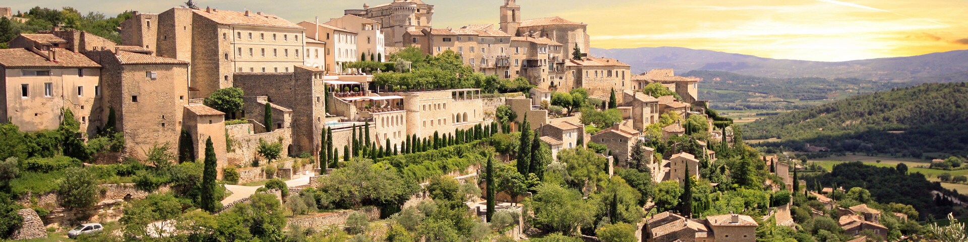 Uma cidade histórica com arquitetura medieval, colinas em socalcos e um pôr do sol ao fundo.