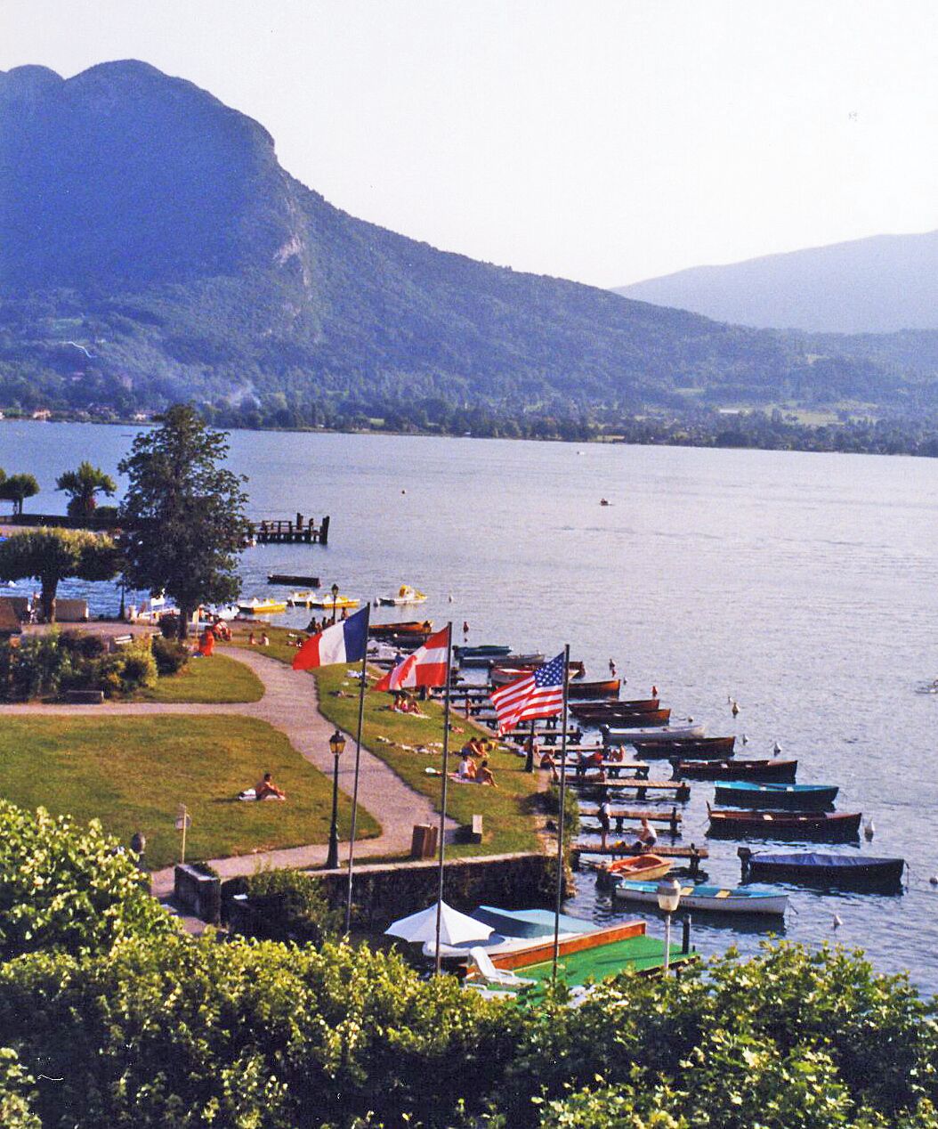 Cena à beira de um lago com barcos atracados na margem, bandeiras e montanhas ao fundo.