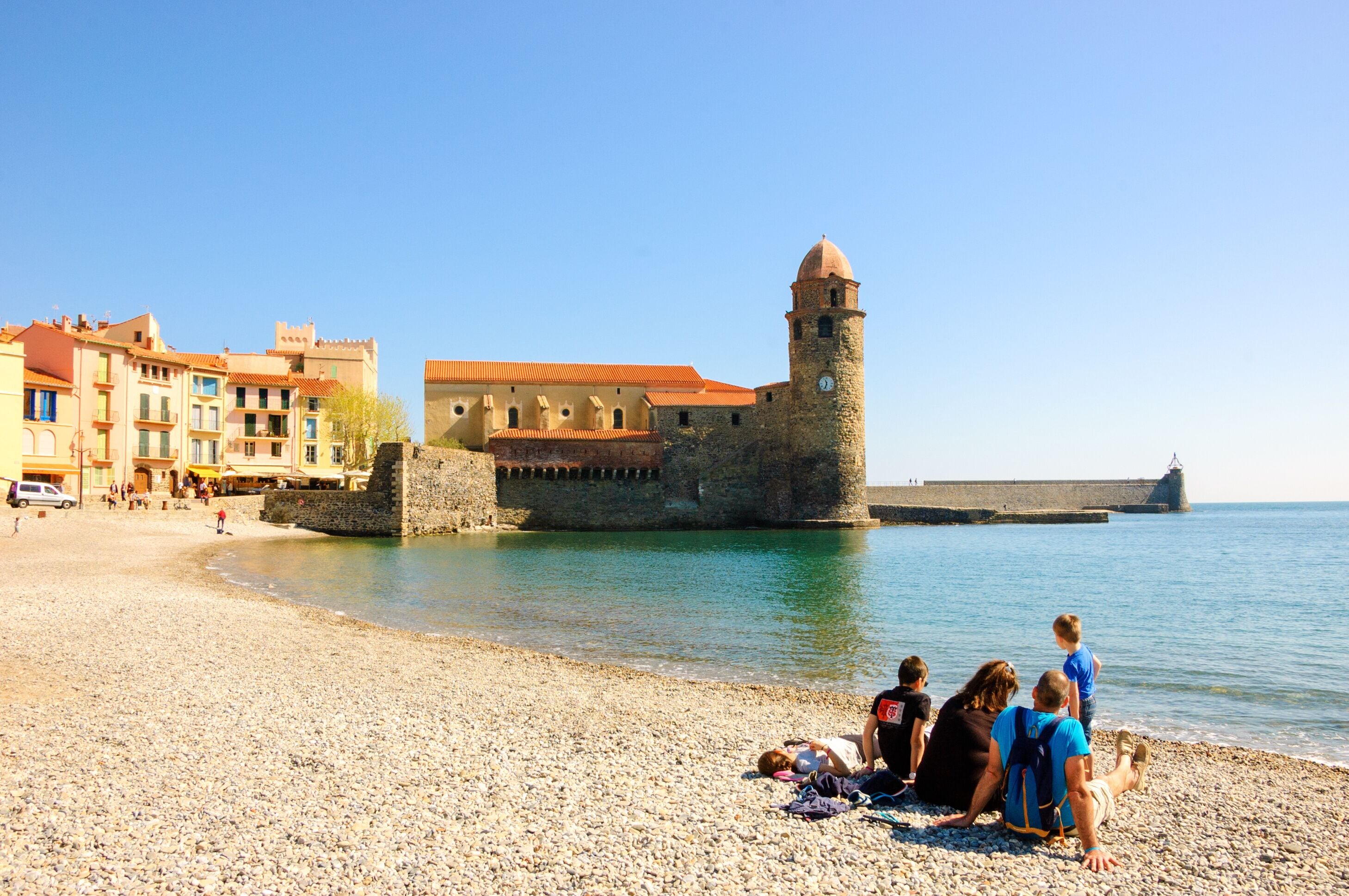 Uma praia de seixos com pessoas relaxando, uma torre de pedra histórica e uma fileira de edifícios coloridos.