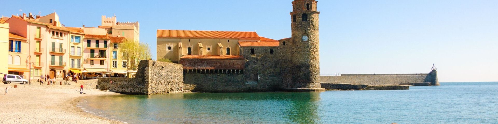 Uma praia de seixos com pessoas relaxando, uma torre de pedra histórica e uma fileira de edifícios coloridos.