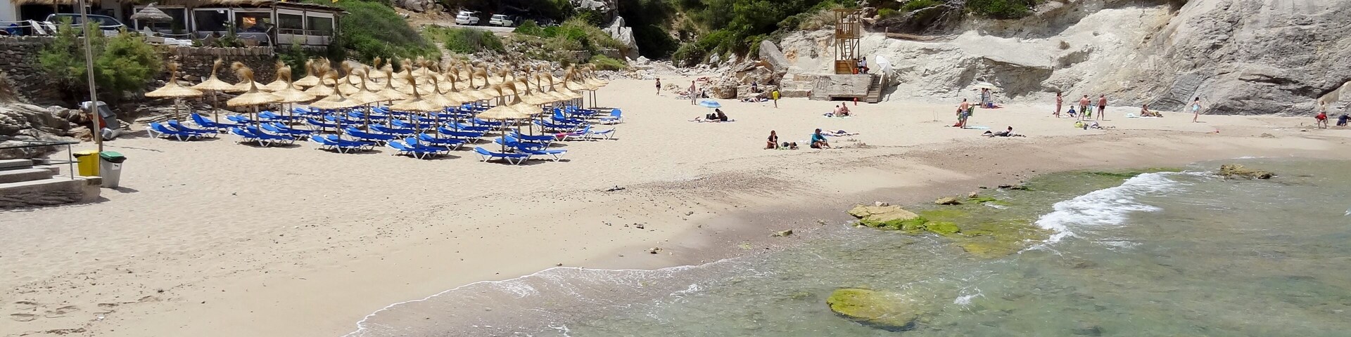 Uma praia com águas cristalinas, guarda-sóis azuis e montanhas rochosas ao fundo.