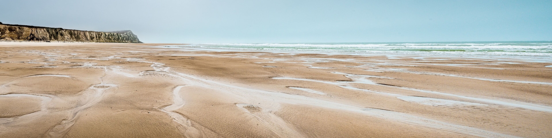 Uma praia de areia com pegadas e um penhasco ao fundo.