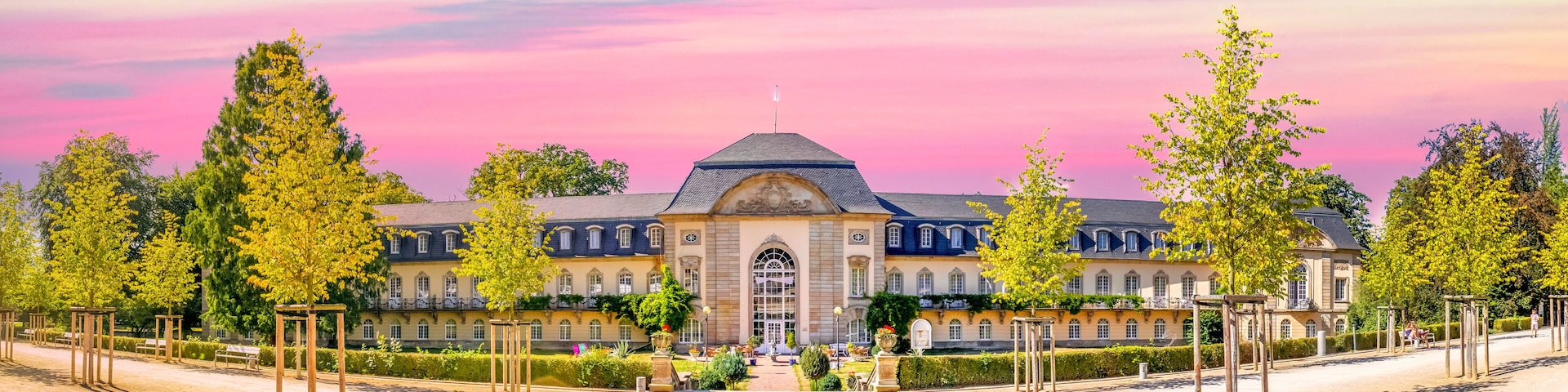 Um edifício histórico com entrada central, ladeado por jardins e árvores simétricas.