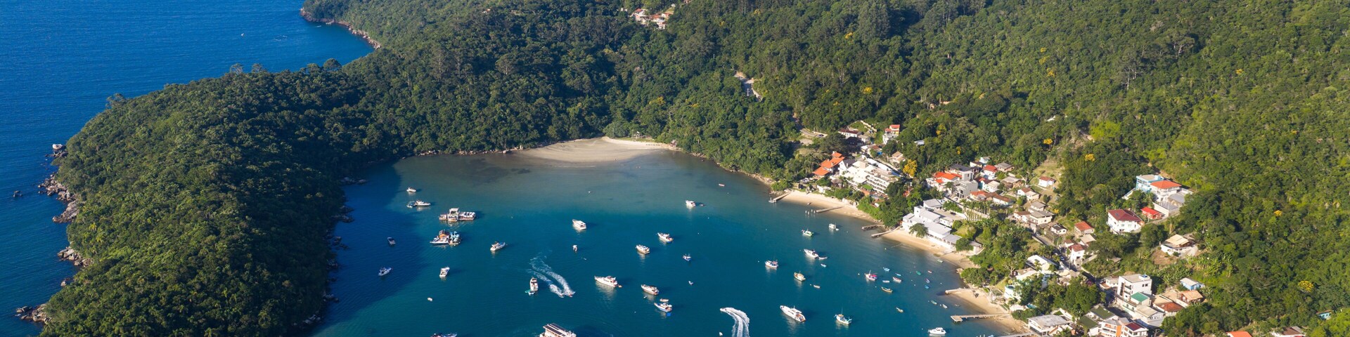 Vista aérea de uma cidade costeira com uma baía, rodeada por floresta densa e colinas.