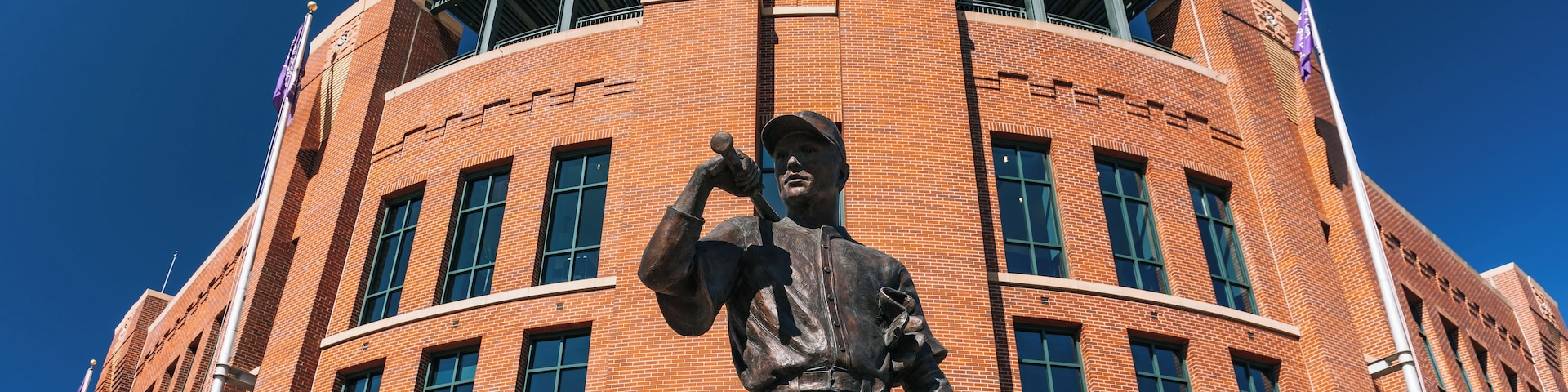 Estátua de um jogador de beisebol em frente a um edifício de tijolos com a inscrição 'COORS FIELD'.