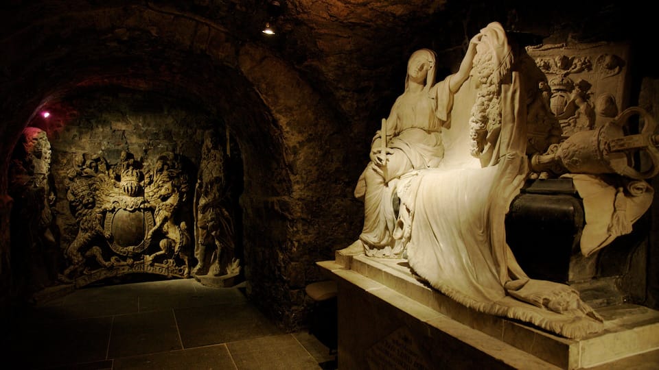 Um sarcófago de pedra com uma figura reclinada, em um túmulo pouco iluminado com um arco de pedra e uma escultura em relevo ao fundo.