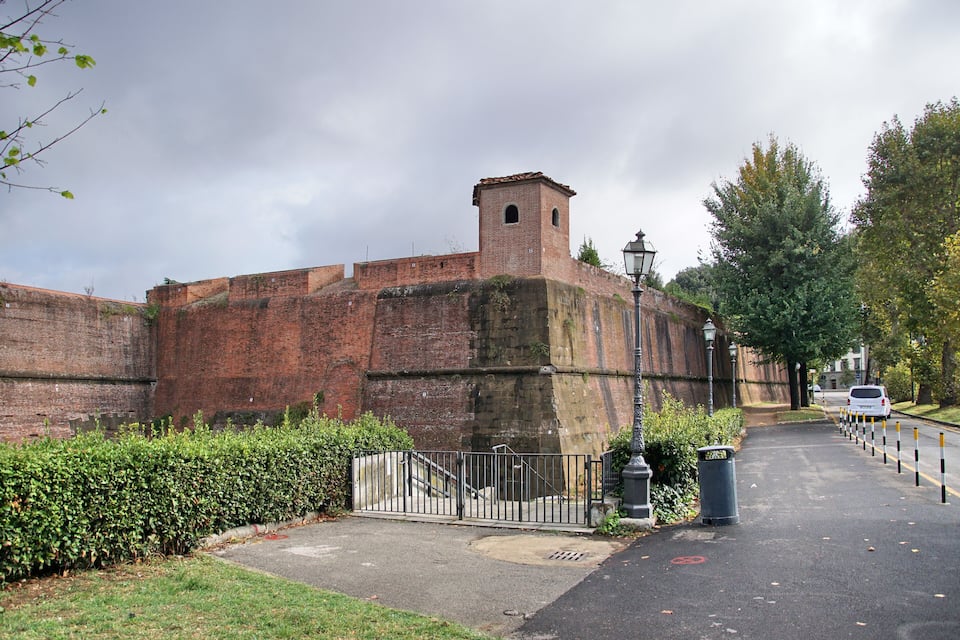 Um muro de tijolos históricos, um caminho pavimentado e uma fileira de defensas ao lado.