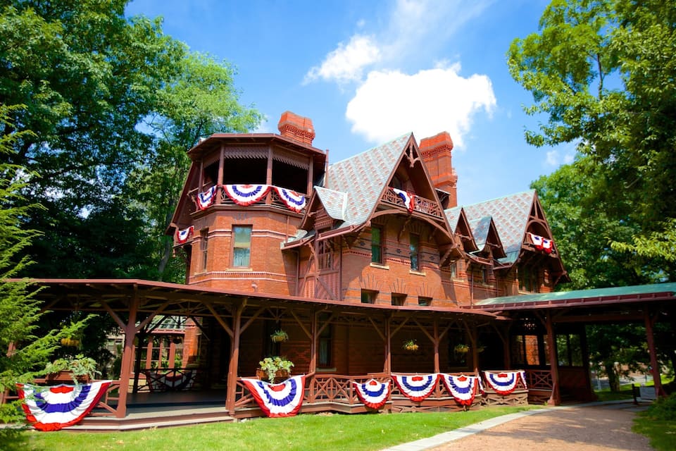 Uma casa grande e ornamentada de tijolos, com varanda que envolve toda a construção, cercada por vegetação exuberante.