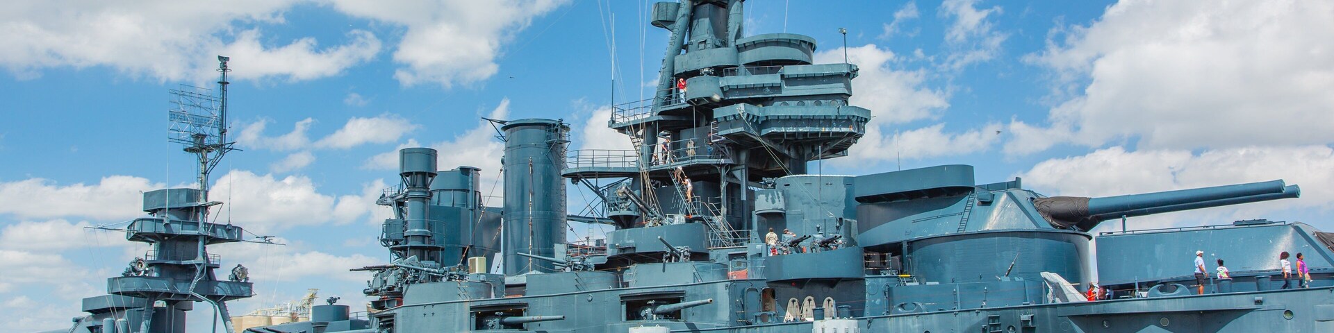 Um grande navio de guerra atracado em um porto, com céu claro e água esverdeada.