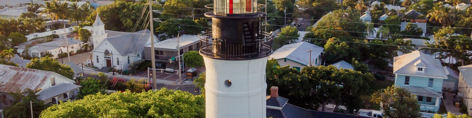 Um farol branco com topo preto, rodeado por vegetação exuberante e edifícios residenciais.