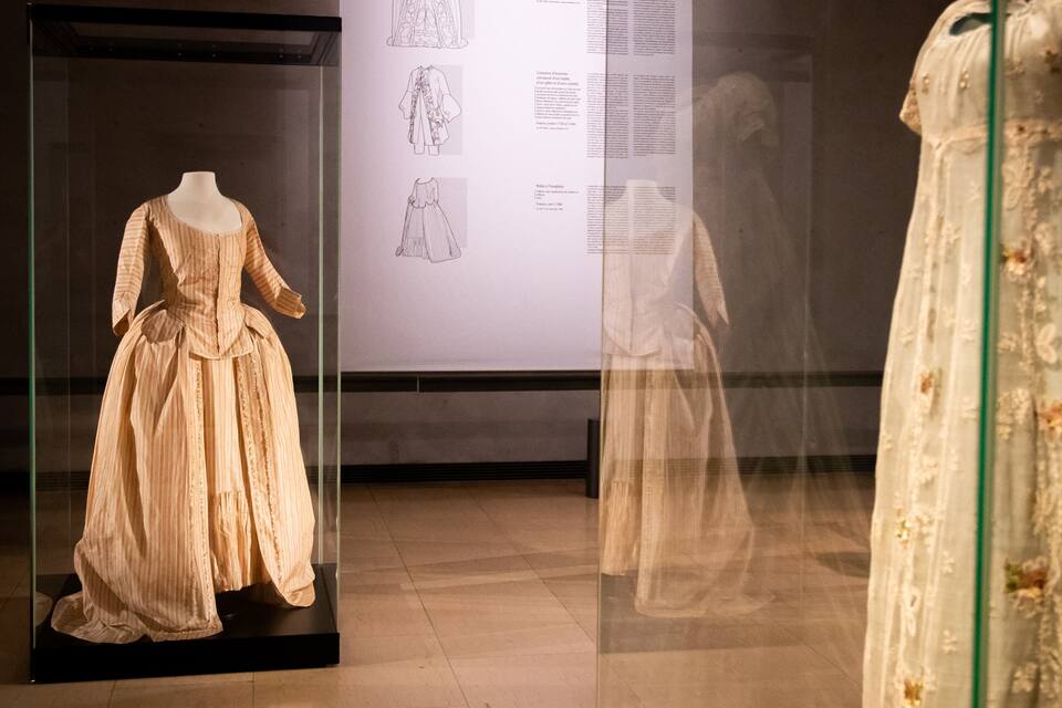 Uma vitrine de museu com um manequim vestindo um vestido listrado e outro de padrão floral.