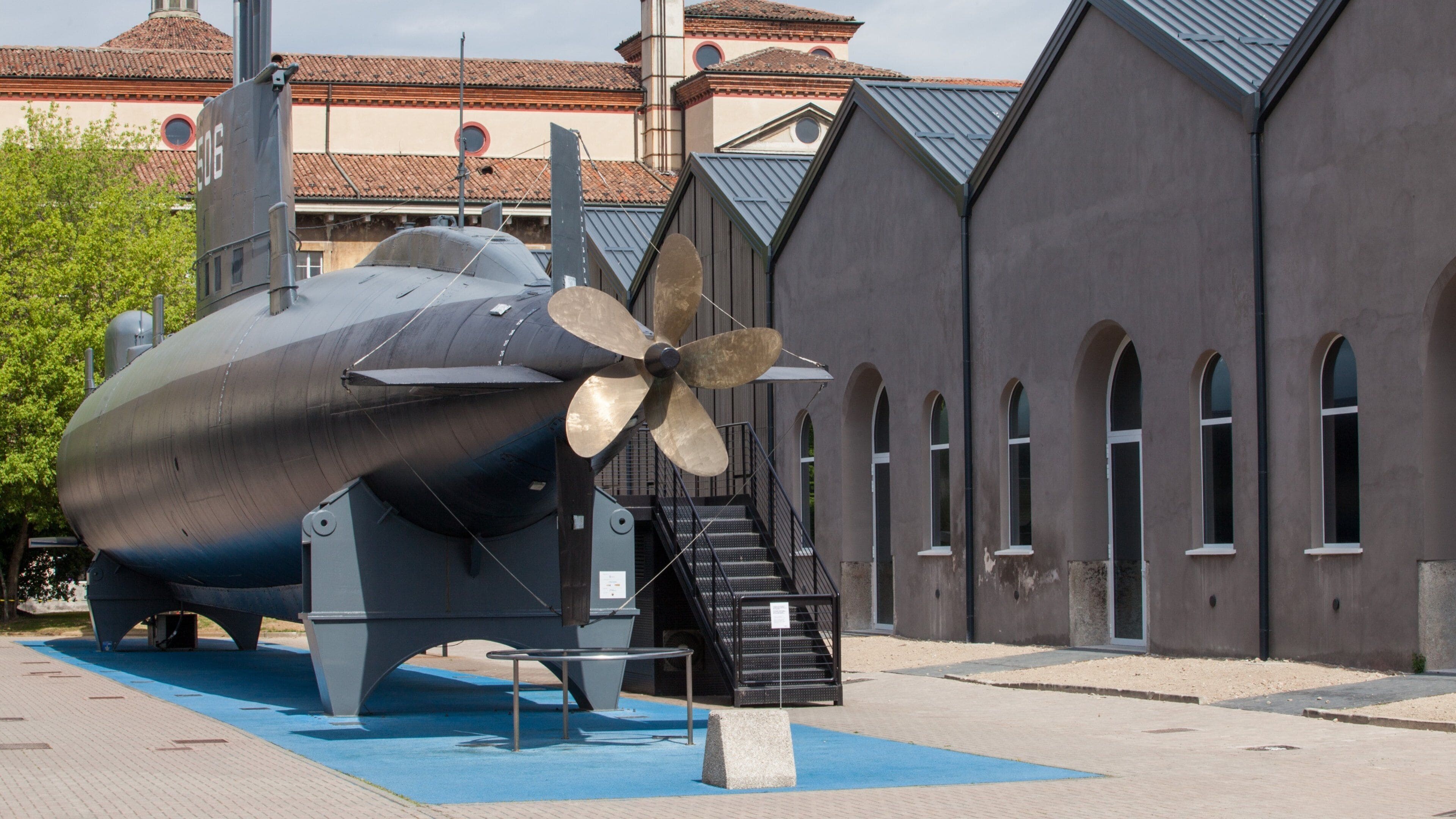 Um submarino em exposição, com uma escadaria que leva até ele, em frente a um edifício com janelas em arco.
