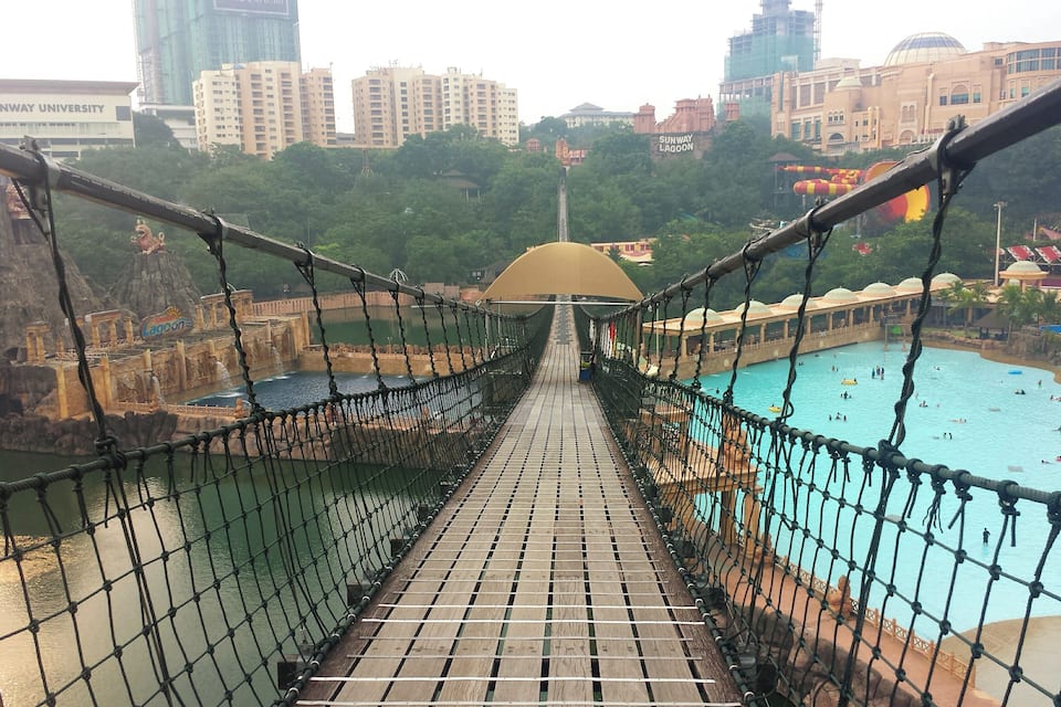 Uma ponte de corda sobre a água, com uma piscina à direita e um edifício grande ao fundo.