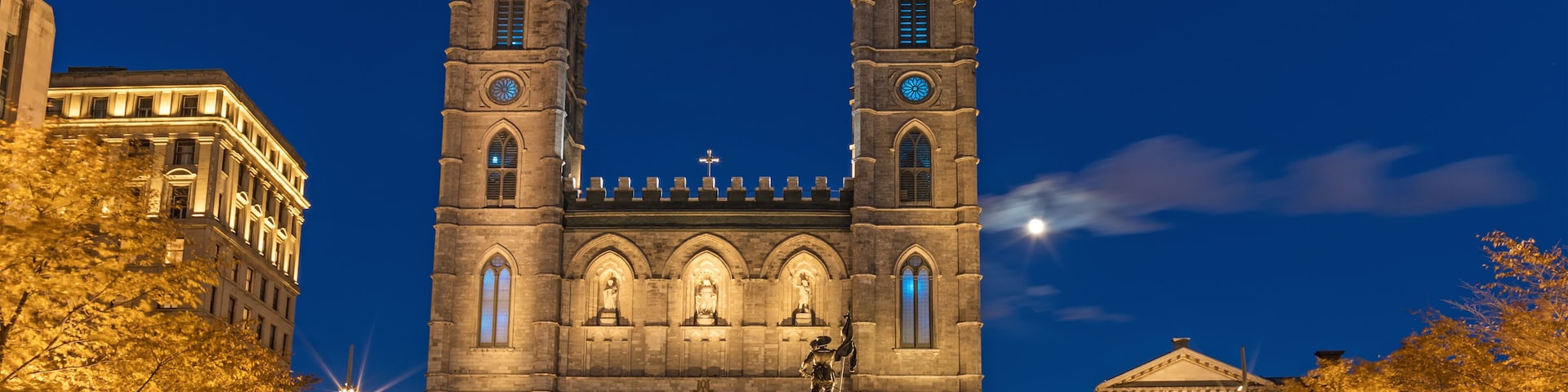 Uma catedral histórica com duas torres e um grande arco central, rodeada por árvores de outono e uma rua movimentada.