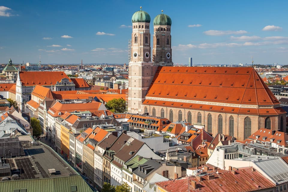 Paisagem urbana com edifícios históricos, uma igreja destacada com duas torres e uma torre moderna ao fundo.