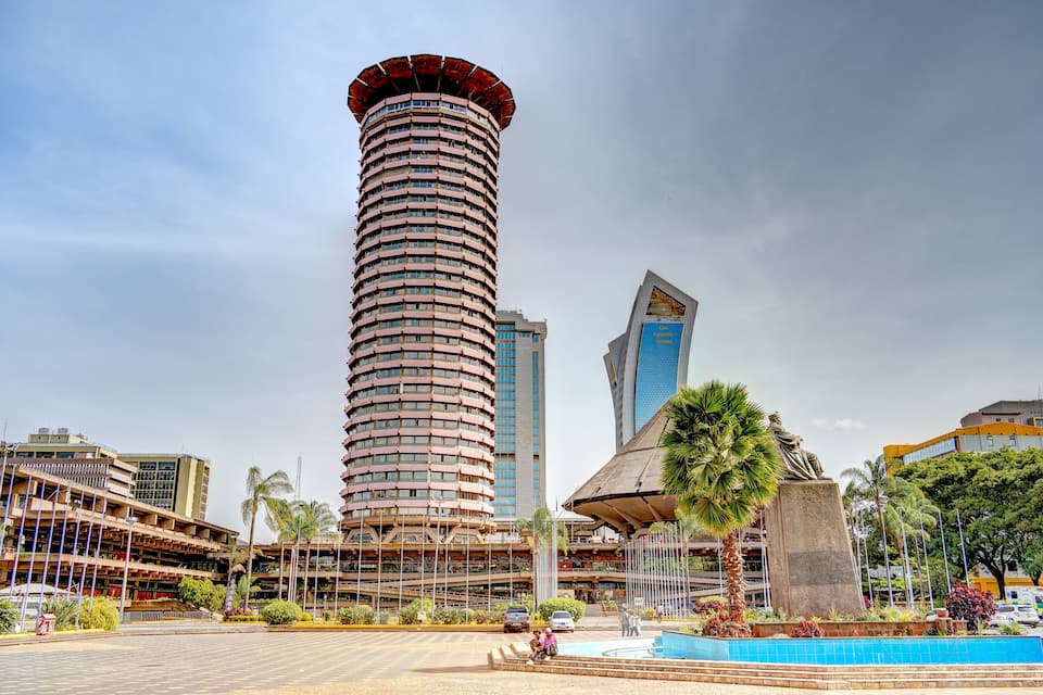 Um edifício cilíndrico alto com telhado vermelho, rodeado por palmeiras e uma estátua.