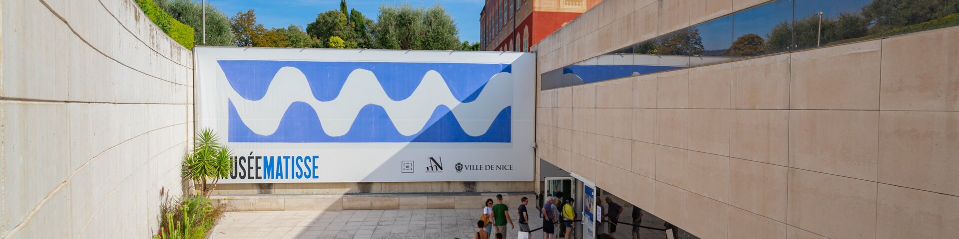 Um edifício moderno com um grande mural azul e branco, um edifício vermelho ao fundo e pessoas caminhando nas escadas.