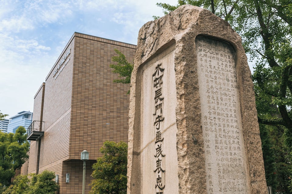 Um monumento de pedra com inscrições, um edifício moderno e árvores ao fundo.