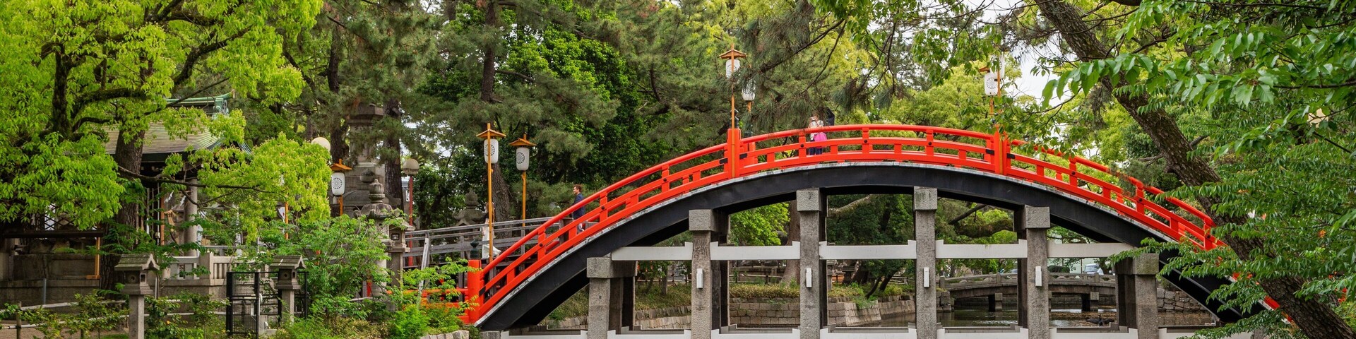 Uma ponte japonesa tradicional sobre um lago com carpas, cercada por vegetação exuberante e arquitetura tradicional.