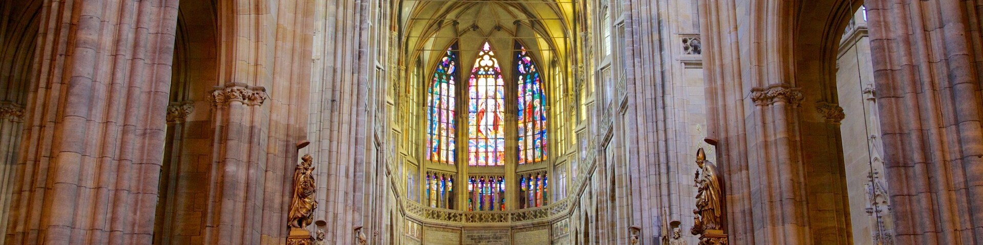 Interior de uma catedral com altos tetos em arco, janelas de vitrais e estátuas ricamente decoradas.