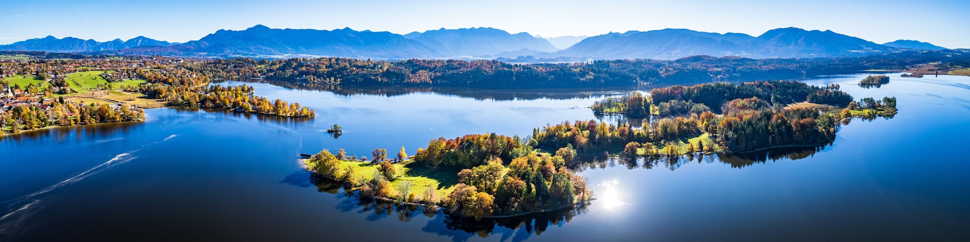 Um lago tranquilo rodeado por árvores em tons de outono e montanhas ao longe.