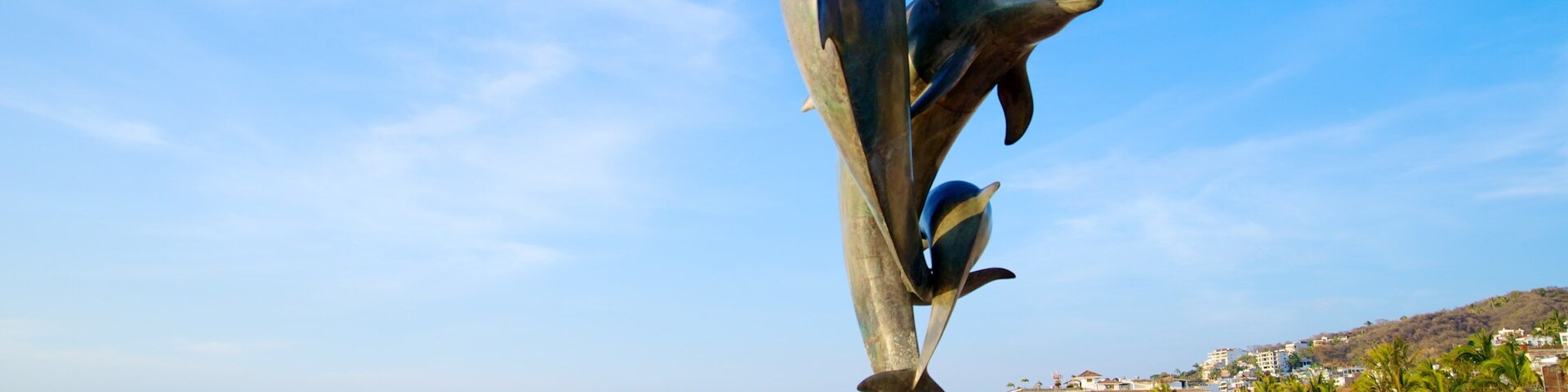 Uma escultura de golfinho numa praia, com uma paisagem urbana ao fundo.