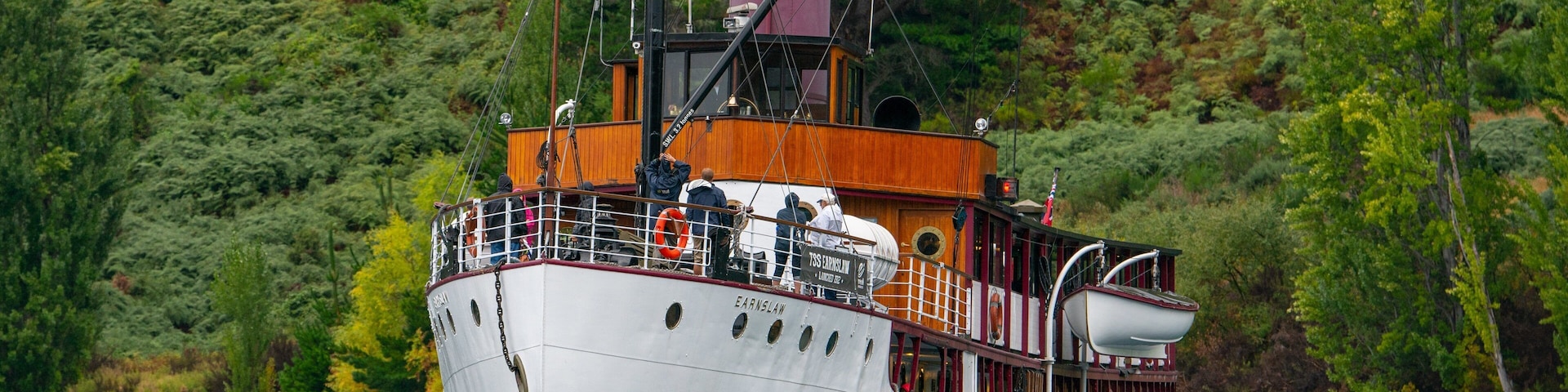 Um barco a vapor branco com faixa vermelha e chaminé, navegando em um rio cercado por floresta verde densa.