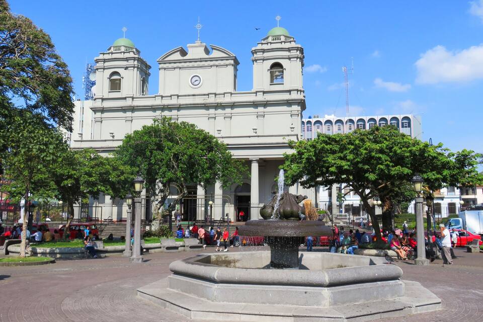 Um edifício histórico com duas cúpulas e uma torre do relógio central, cercado por uma praça pública com uma fonte e árvores.