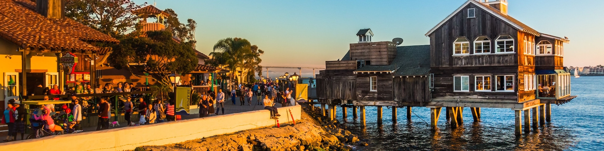 Um calçadão à beira-mar com edifícios de madeira, um litoral rochoso e céu claro.