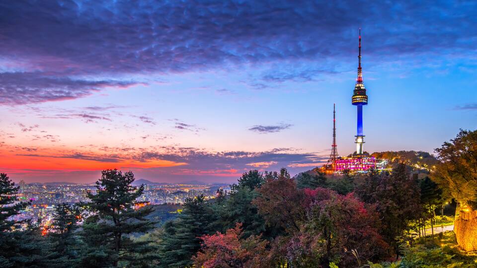 Paisagem urbana ao anoitecer, com uma torre imponente, árvores densas e um céu com tons de pôr do sol.