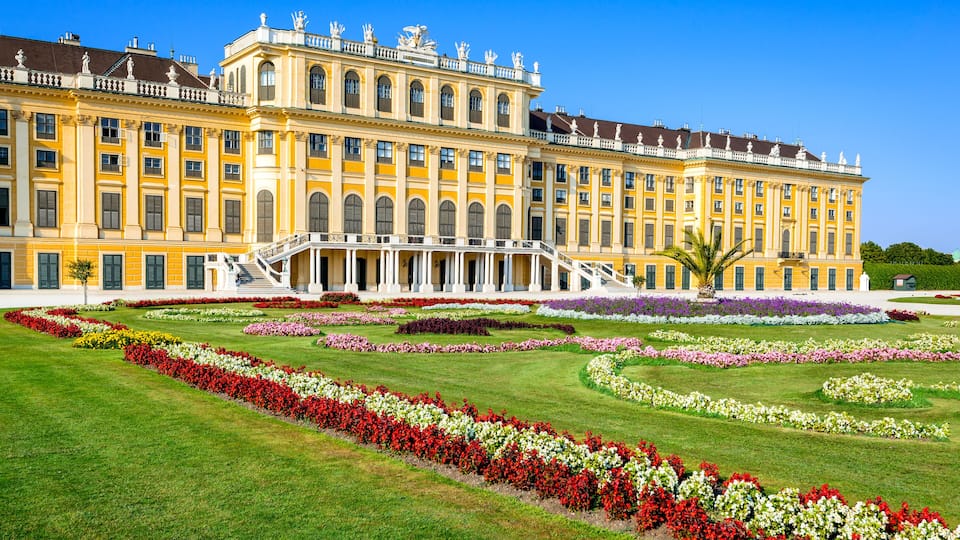 Um edifício grande e ornamentado, com fachada simétrica, várias varandas e um jardim bem cuidado, com diversos canteiros de flores.