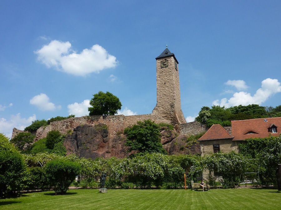 Uma torre histórica com relógio, cercada por vegetação exuberante e um gramado bem cuidado.