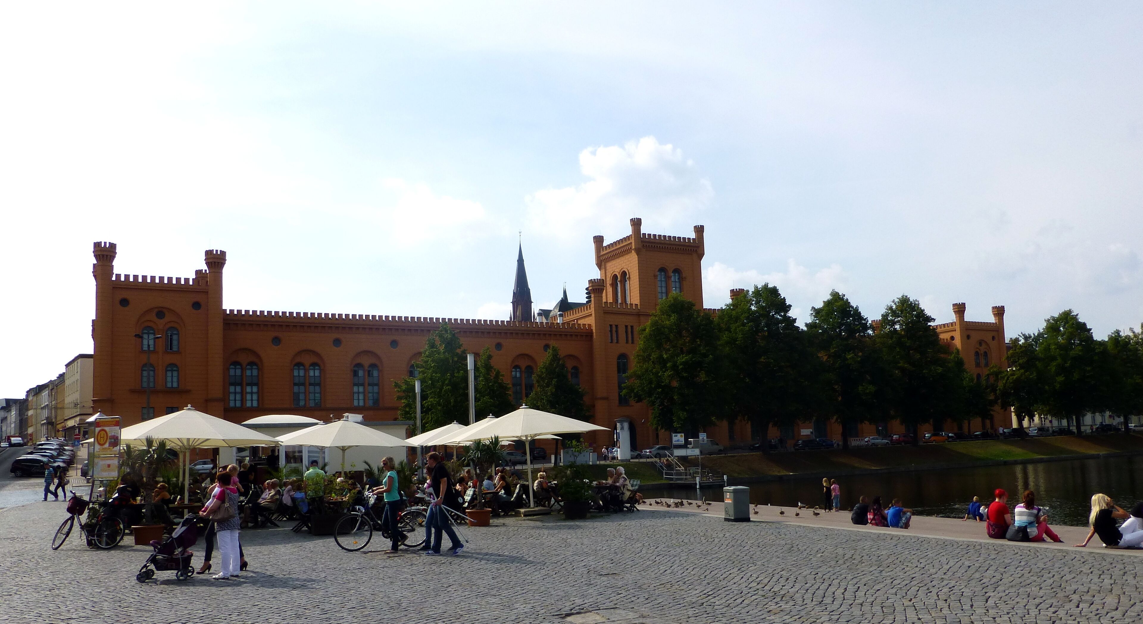 Um edifício histórico com fachada semelhante a um castelo, uma praça de calçamento e um rio ao fundo.