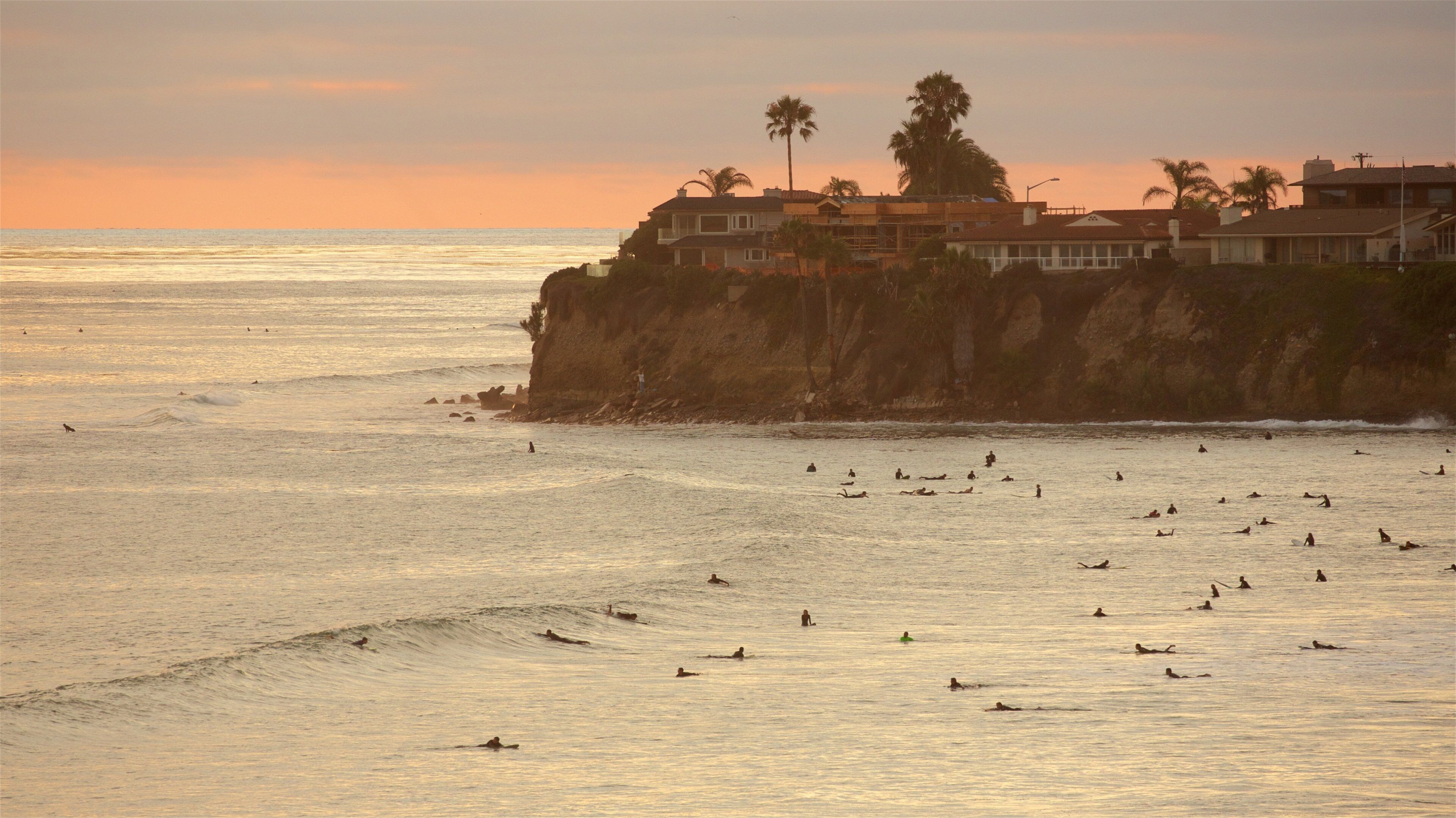Cena costeira com surfistas na água, um pôr do sol e edifícios em um penhasco.