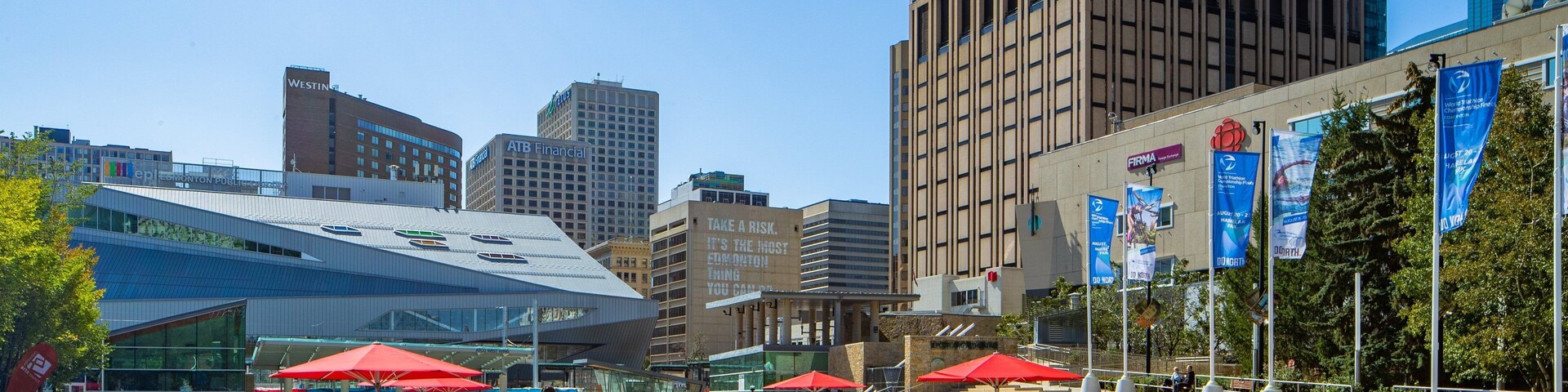 Uma praça urbana com edifícios modernos, guarda-sóis vermelhos e pessoas.
