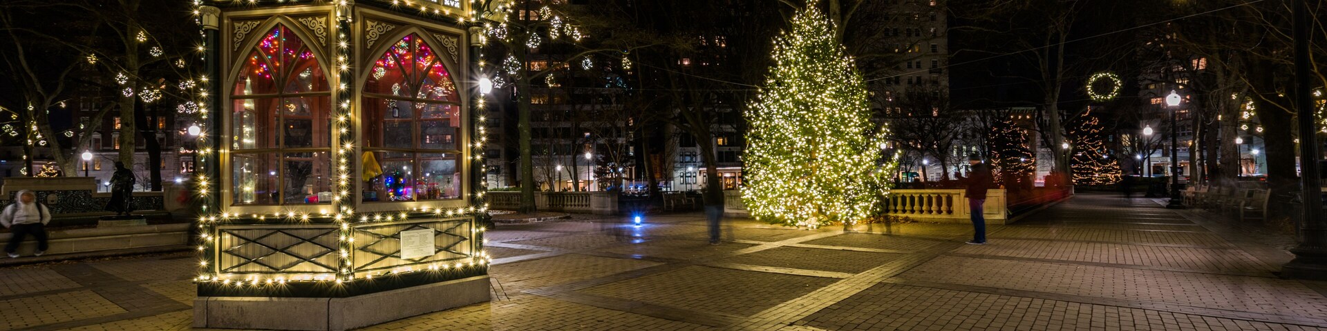 Um pavilhão bem iluminado com desenhos elaborados, uma grande árvore de Natal enfeitada com luzes e uma atmosfera festiva em uma praça da cidade à noite.