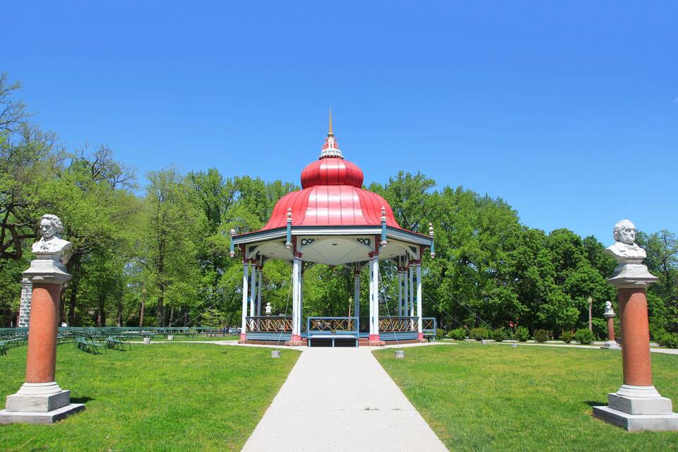 Um gazebo vermelho com colunas brancas e estátuas em um parque.