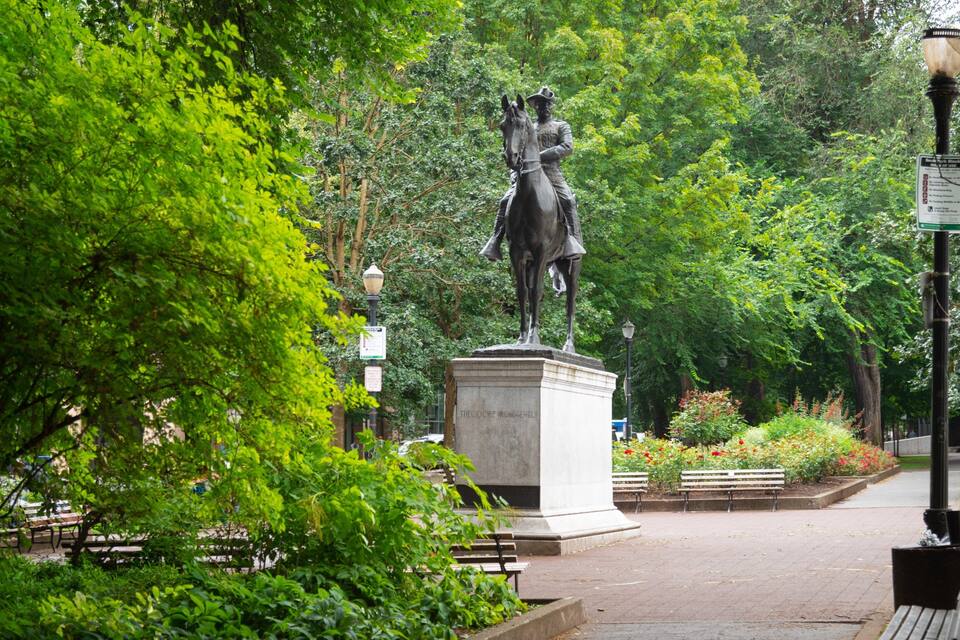 Estátua de Theodore Roosevelt em um parque com bancos, árvores e um lampião.