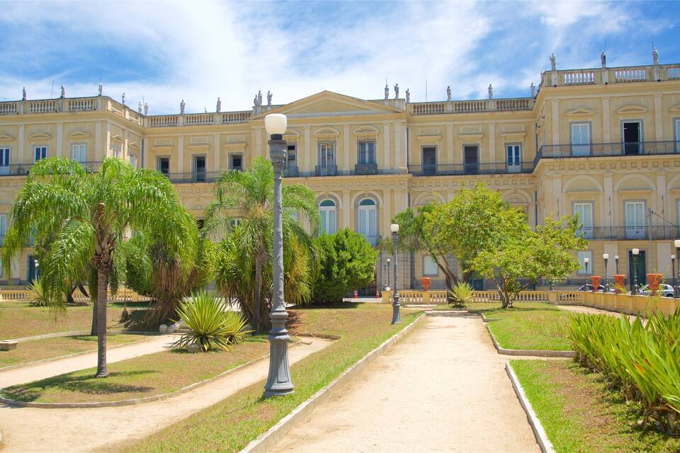 Um edifício imponente com arquitetura clássica, rodeado por jardins bem cuidados e palmeiras.