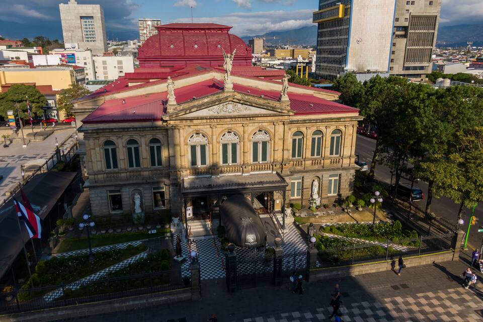 Um edifício histórico com telhado vermelho e arquitetura clássica, rodeado por uma praça com estátuas e uma fonte.