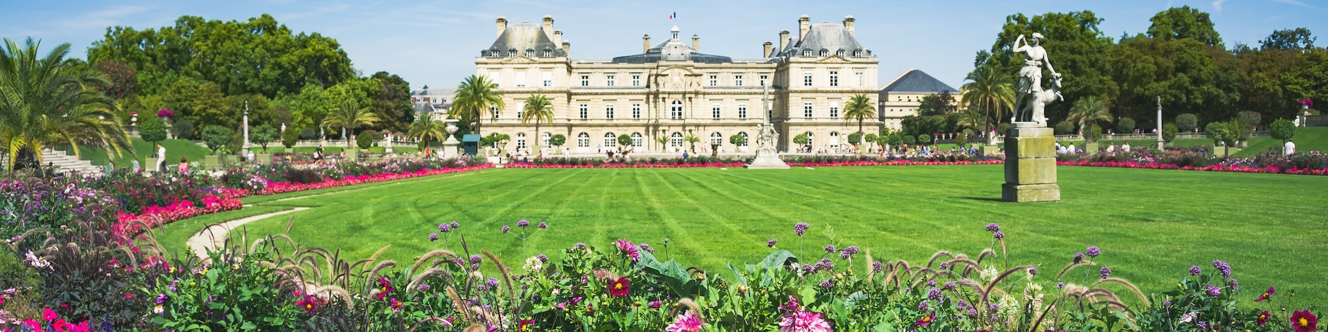 Um castelo imponente com um jardim bem cuidado, canteiros de flores vibrantes e uma estátua em primeiro plano.