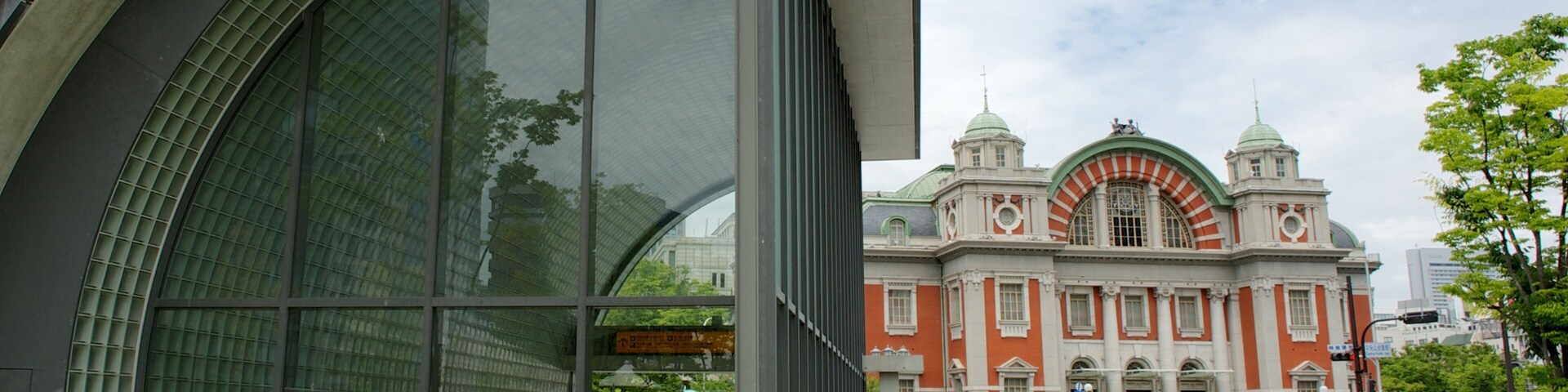 Um edifício moderno de vidro com fachada curva, um edifício histórico vermelho com cúpulas ao fundo, e um céu claro.