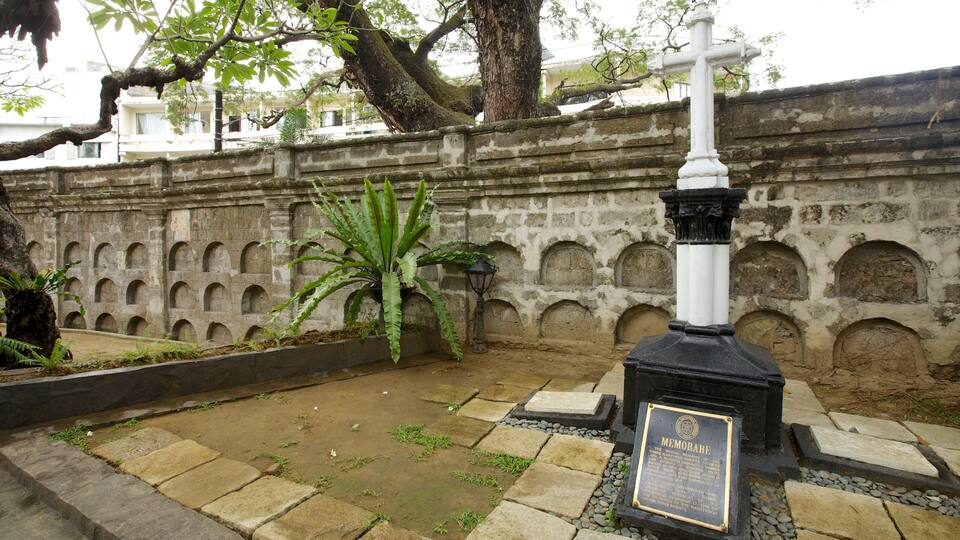 Um memorial com uma cruz, uma placa e um muro de pedra com arcos.