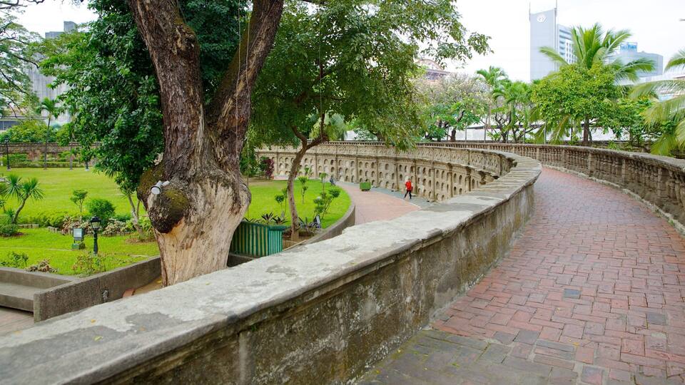 Um muro de pedra curvo com um caminho de tijolos, rodeado por vegetação exuberante e árvores.