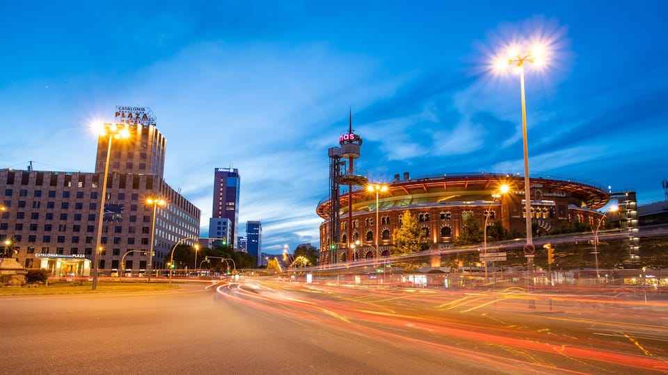 Paisagem urbana com um edifício circular e uma torre do relógio, rodeado por edifícios modernos e um cruzamento movimentado com rastros de luz.