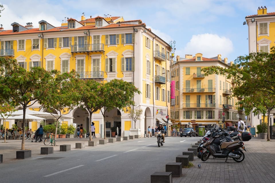 Rua com edifícios amarelos, árvores e motocicletas estacionadas.