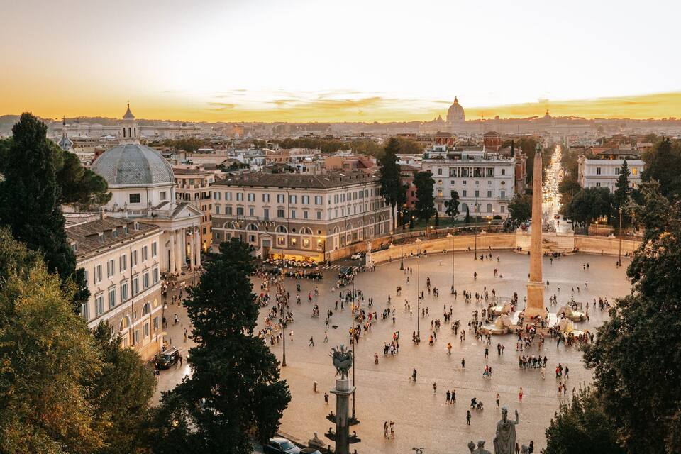 Uma praça movimentada com edifícios históricos, uma cúpula imponente e um monumento central.