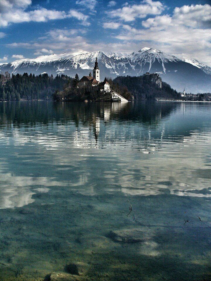 Um lago tranquilo com uma igreja numa ilha, rodeado por montanhas cobertas de neve.
