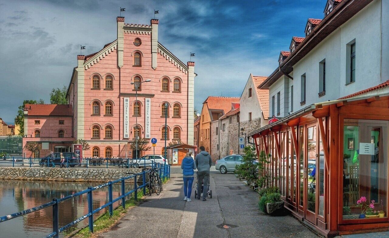 Um edifício histórico com janelas em arco e fachada vermelha, um canal com guarda-corpo azul e um café com toldo vermelho.